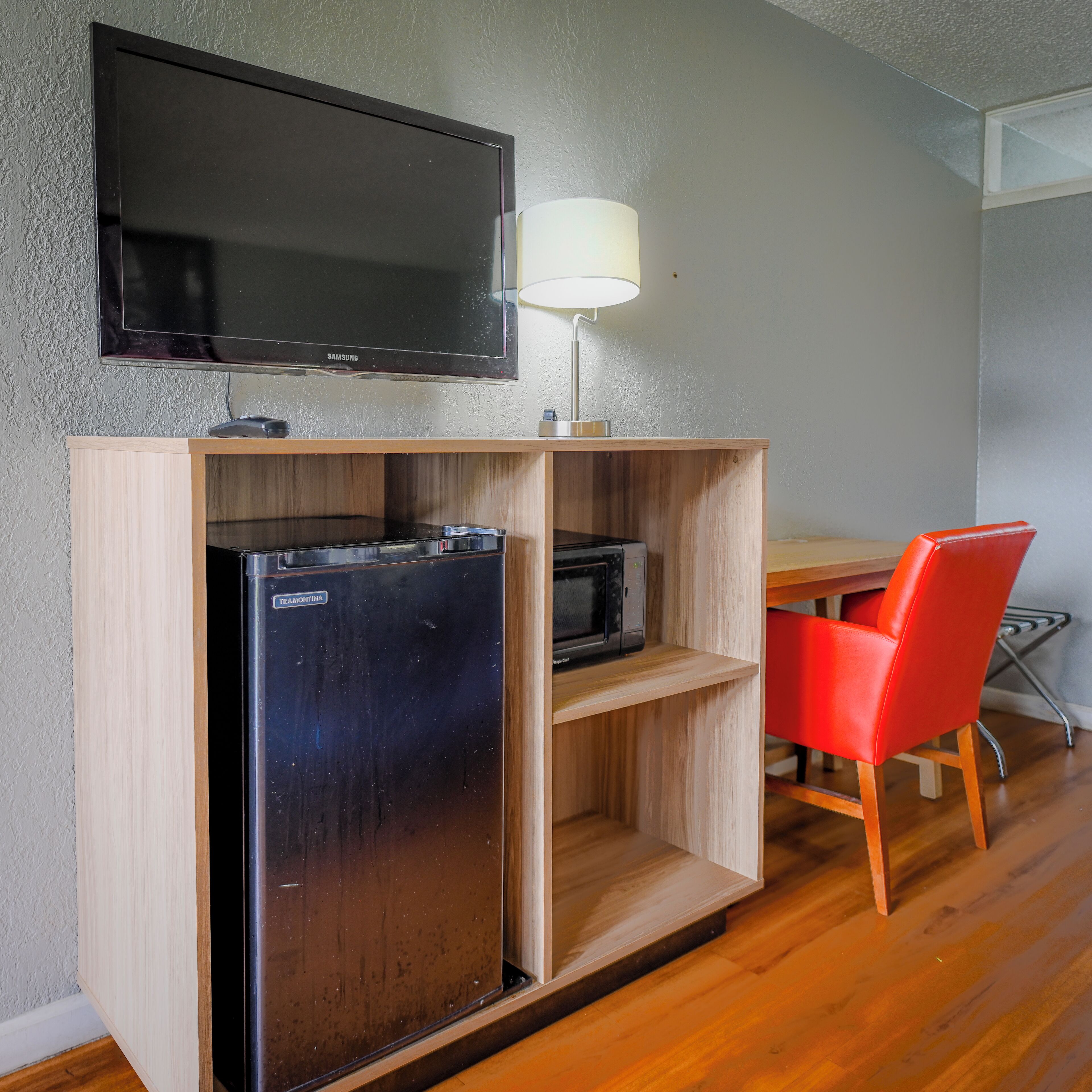 Quarto compacto com televisão de tela plana na parede, mini-frigorífico, mesa e uma cadeira vermelha.