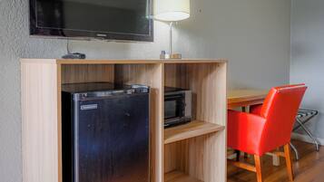 Quarto compacto com televisão de tela plana na parede, mini-frigorífico, mesa e uma cadeira vermelha.
