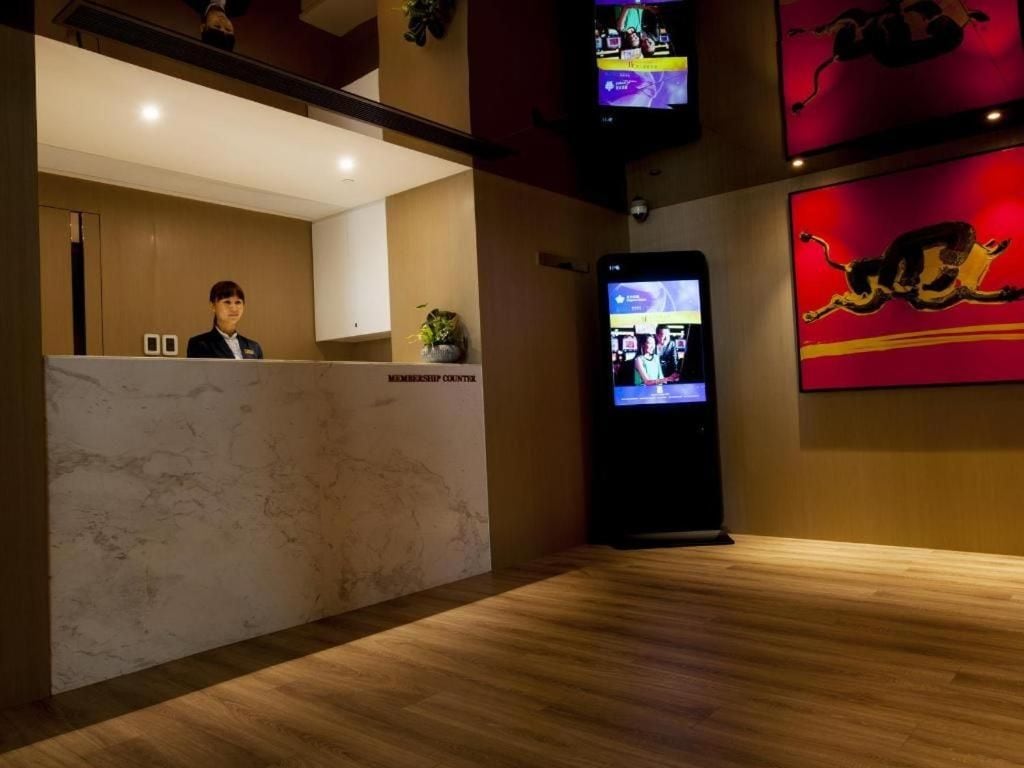 Área de recepção de hotel com balcão, quiosque digital e televisão na parede.