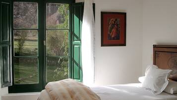 Quarto com cama, janela com persianas verdes, quadro na parede e vista para o verde lá fora.
