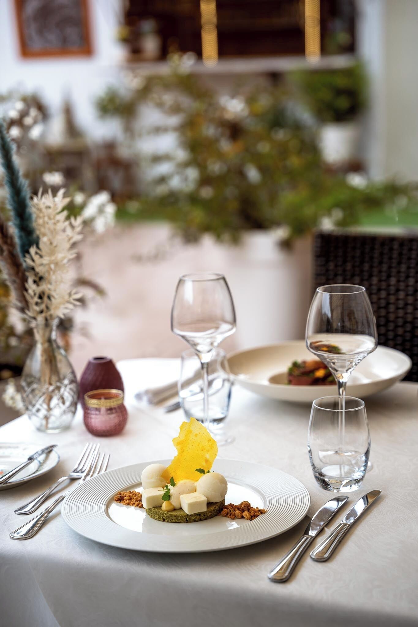 Mesa de jantar posta com pratos, talheres e copos, com um vaso decorativo e uma planta ao fundo.