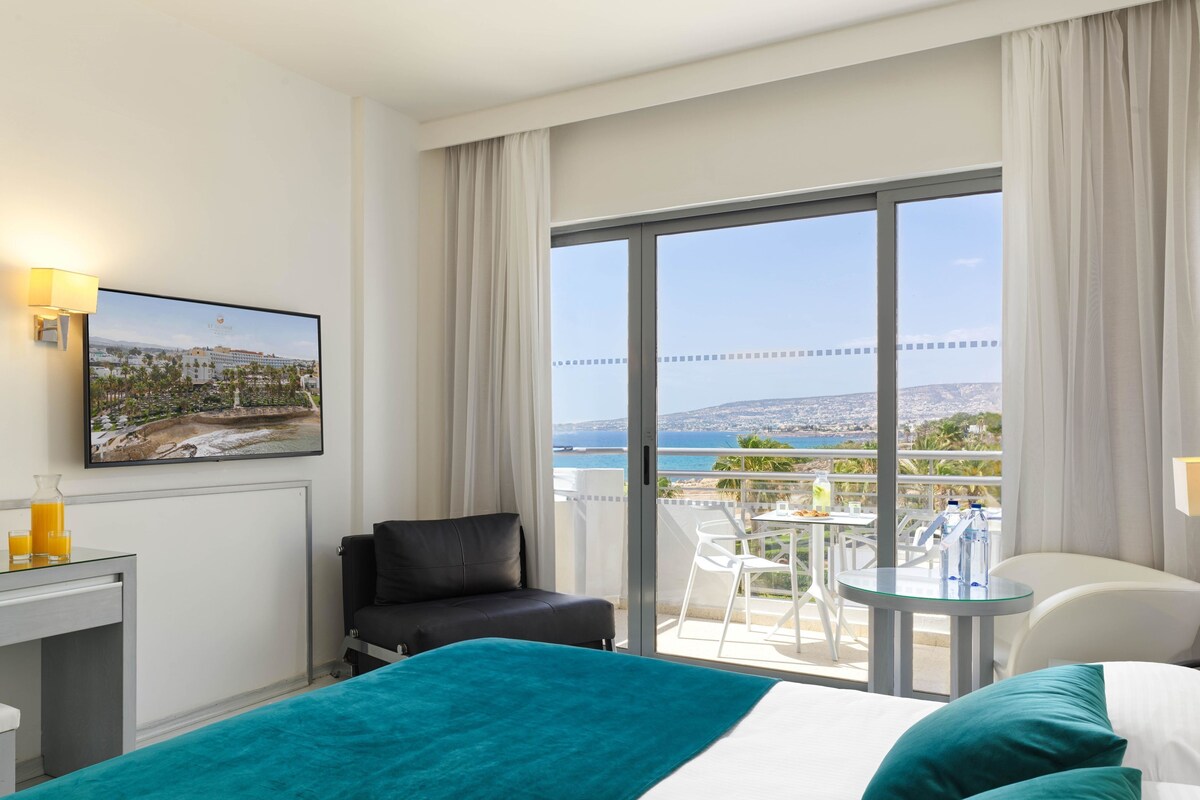 Quarto de hotel com cama, escrivaninha com abajur, cadeira e varanda com mesa e cadeiras.