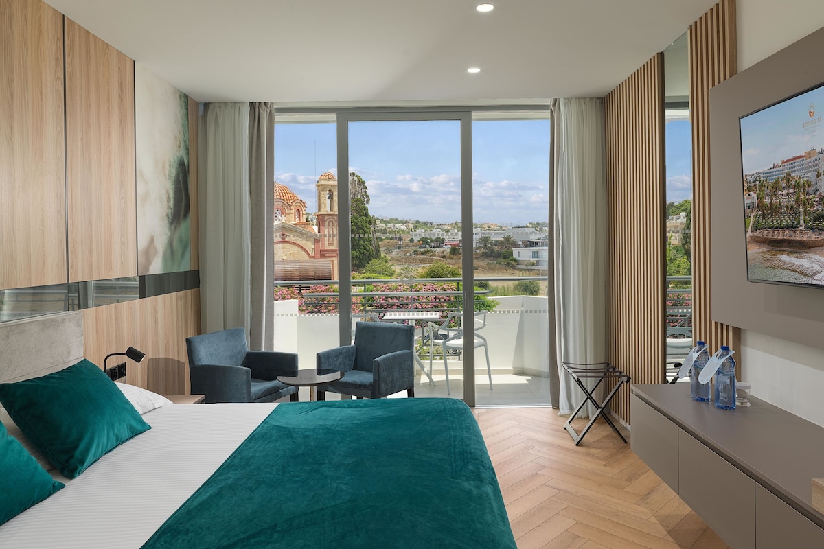Quarto de hotel com cama, duas cadeiras, uma mesinha, TV e varanda com vista para a cidade e um edifício.