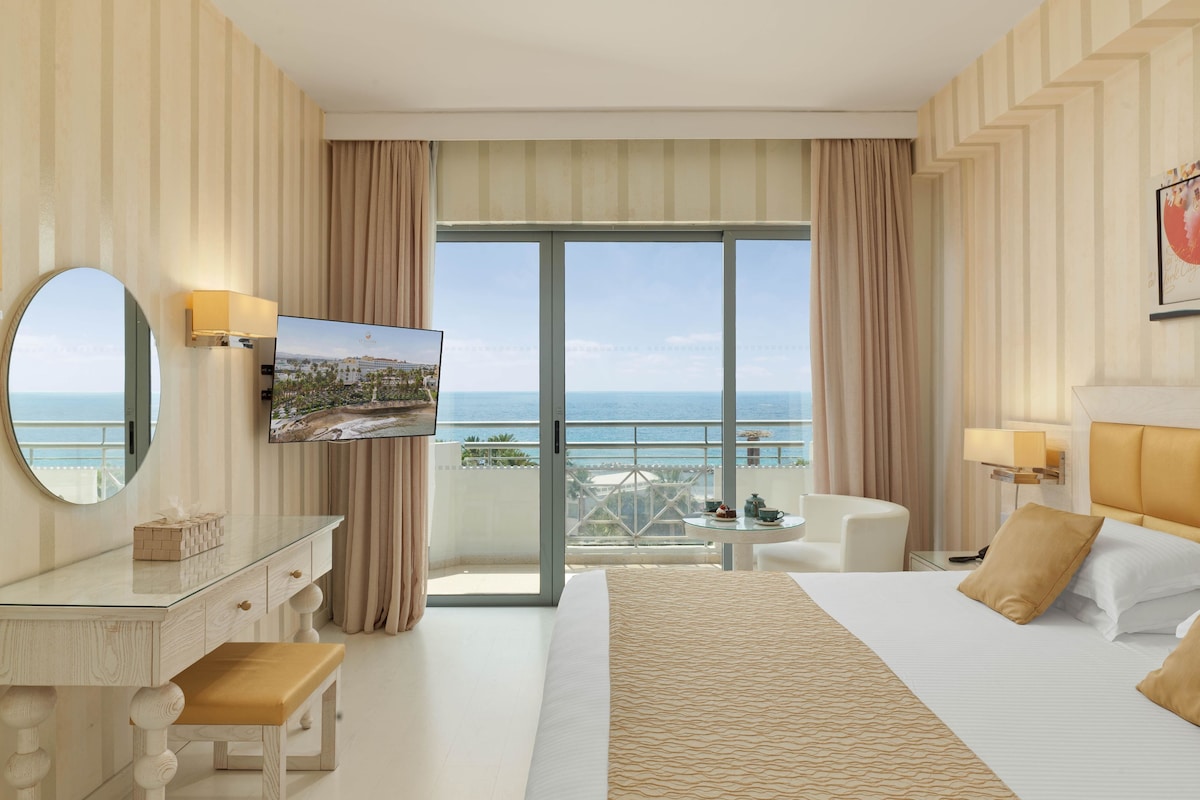 Quarto de hotel com uma cama grande, uma mesa de escritório com espelho, televisão e varanda com vista para o mar.