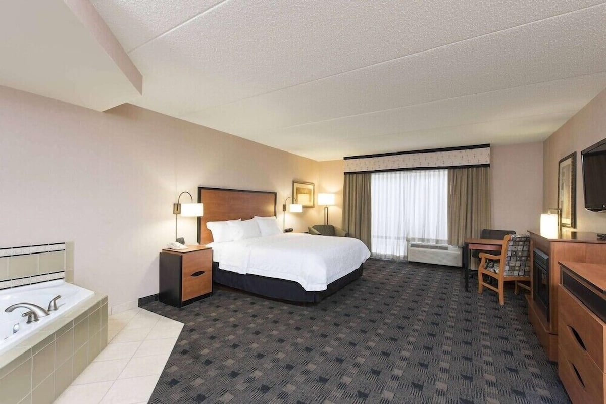 Quarto de hotel com cama, escrivaninha, cadeira, televisão, luminária, mesa de cabeceira e banheiro com banheira.
