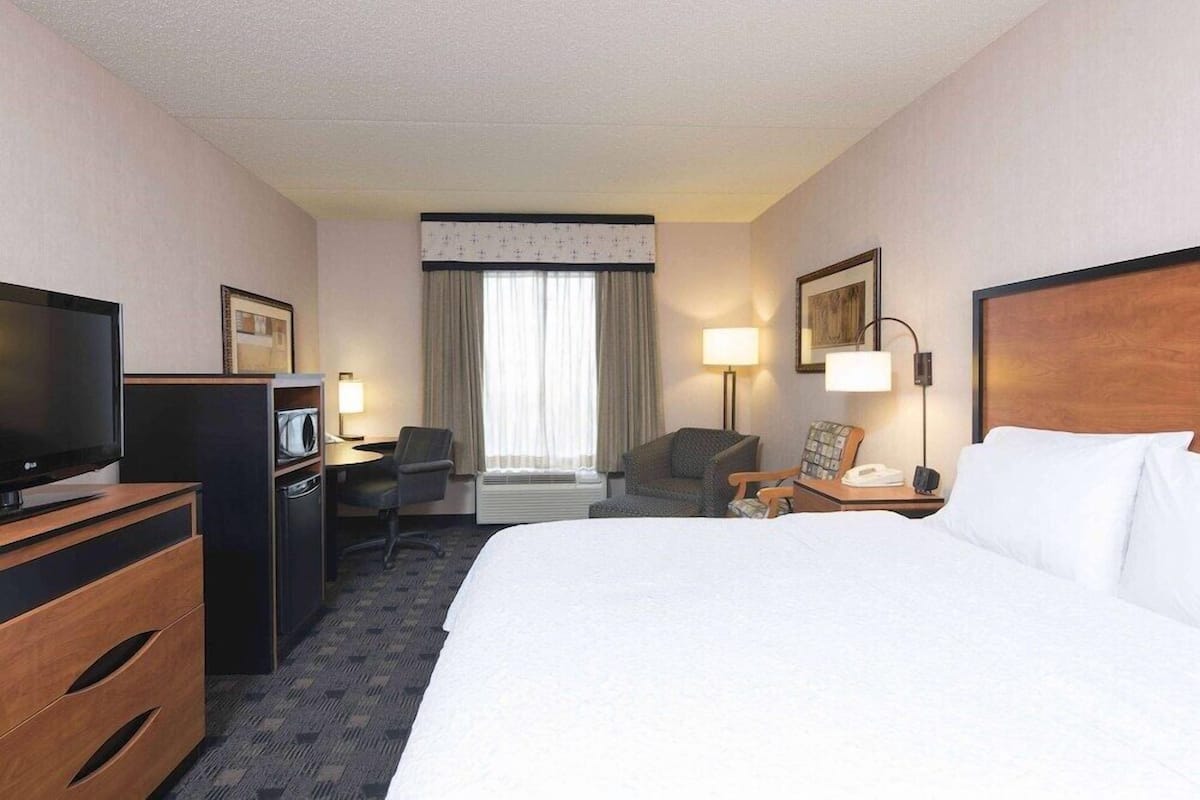 Quarto de hotel com cama, escrivaninha, televisão, cadeira, abajur e mesa de cabeceira com telefone.
