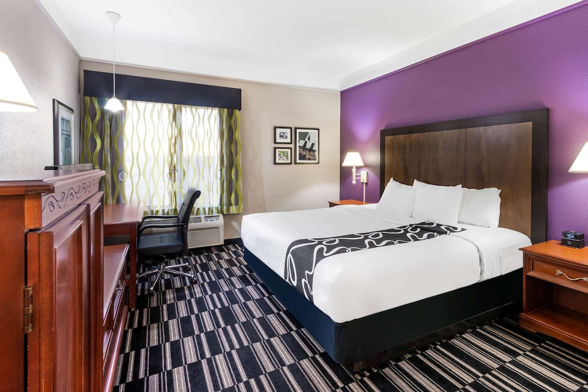 Quarto de hotel com uma cama grande, uma escrivaninha com cadeira, uma luminária de mesa e um relógio fixado na parede.