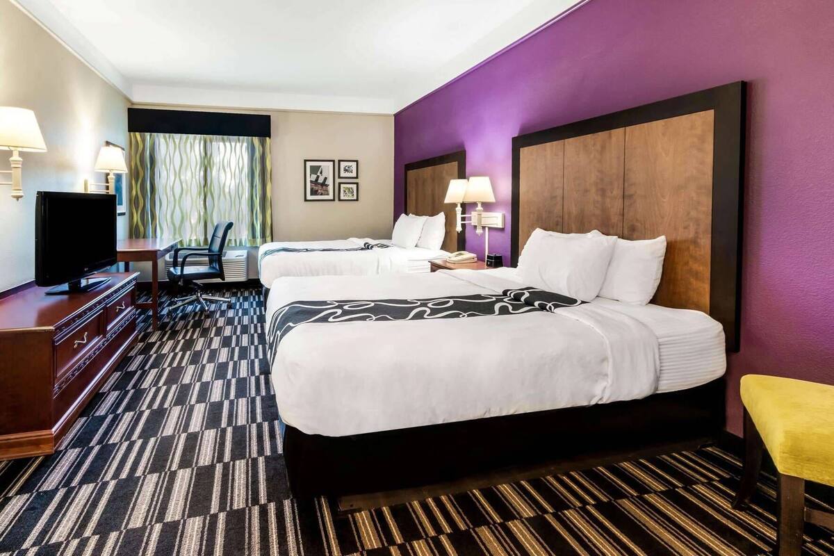 Quarto de hotel com duas camas, uma escrivaninha com televisão, uma cadeira, um abajur e um tapete estampado.