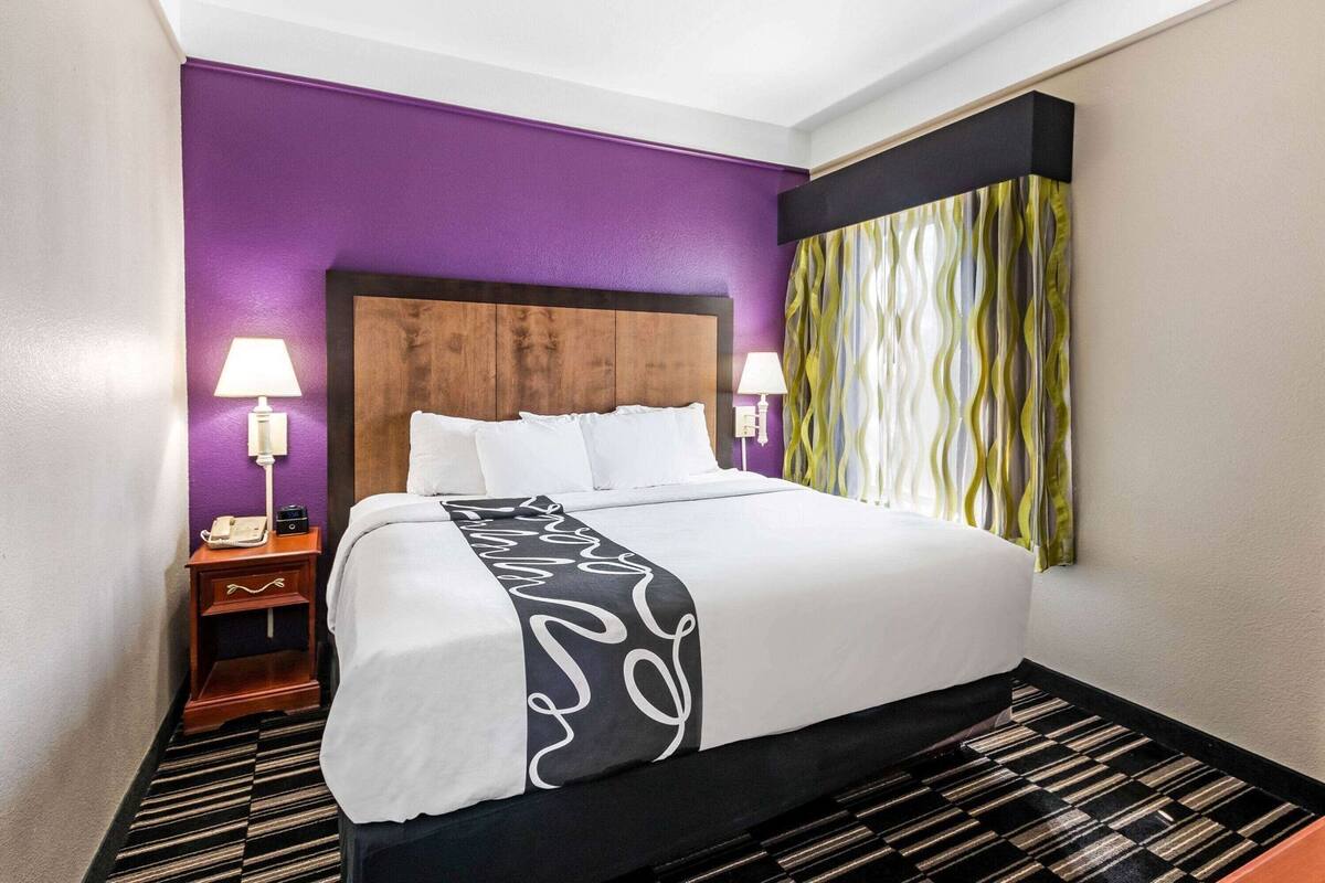 Quarto de hotel com uma cama, duas mesas de cabeceira, um abajur, um telefone e cortina estampada.