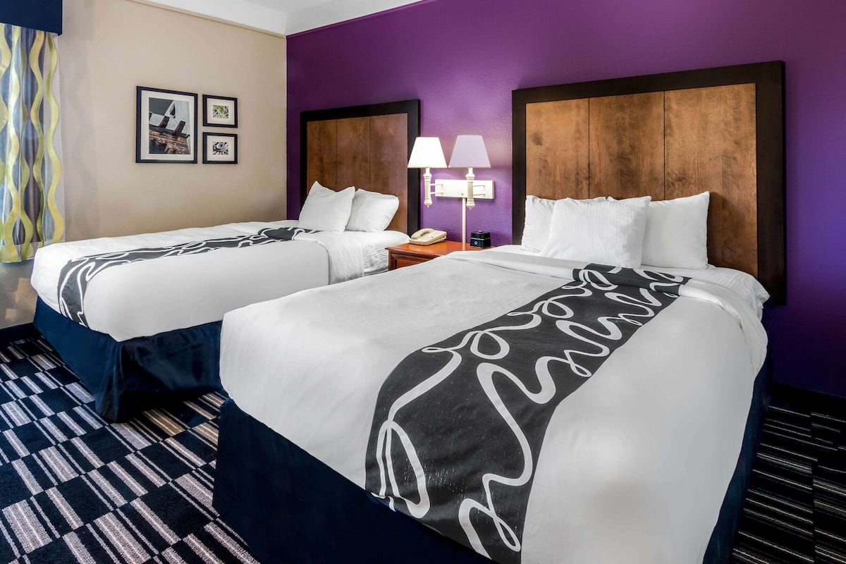 Quarto de hotel com duas camas, uma mesinha de cabeceira, uma luminária e quadros nas paredes.