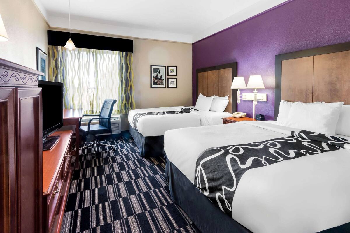 Quarto de hotel com duas camas, uma escrivaninha com televisão, uma cadeira e uma janela com cortinas.