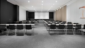 Uma sala de conferência com fileiras de mesas e cadeiras, um púlpito e uma tela grande na frente.