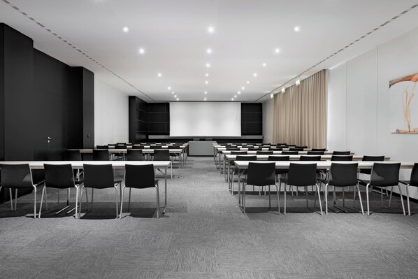 Uma sala de conferência com fileiras de mesas e cadeiras, um púlpito e uma tela grande na frente.