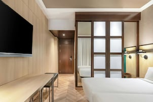 Quarto de hotel com cama, escrivaninha, televisão e porta deslizante para um banheiro.