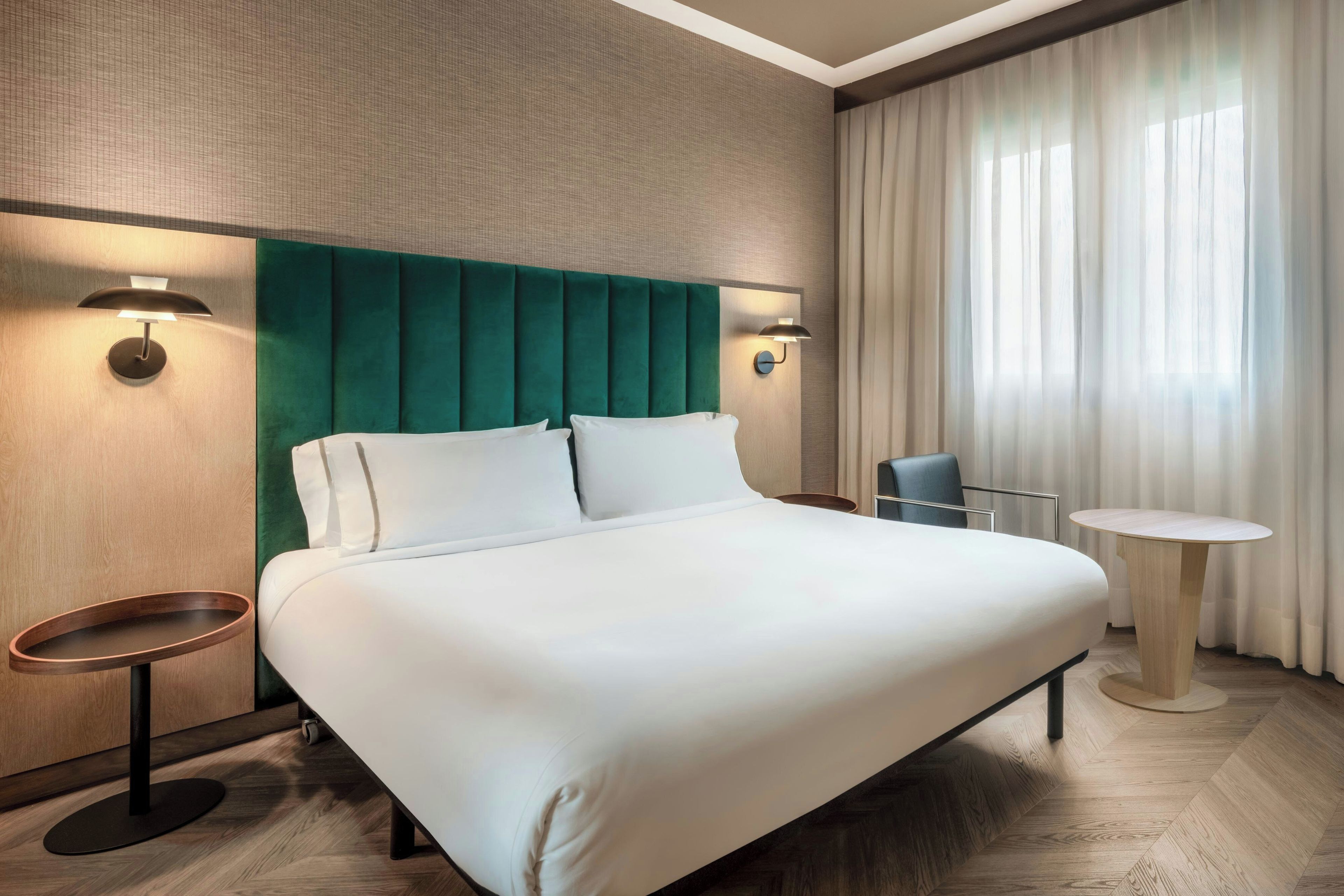 Quarto de hotel com cama grande, duas mesinhas de cabeceira, uma cadeira, uma mesinha e uma janela com cortinas.