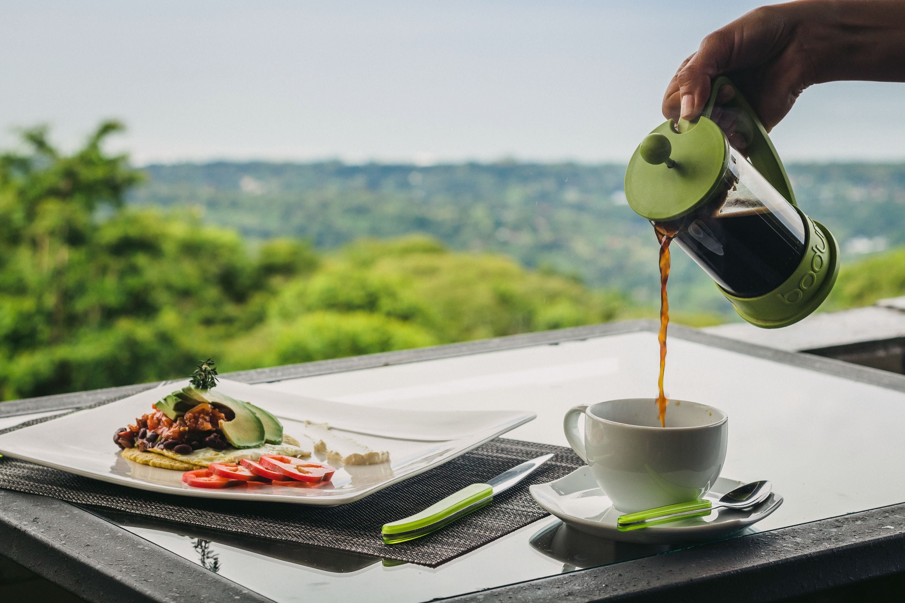 Uma mão servindo café em uma xícara sobre uma mesa com um prato de comida e uma faca.