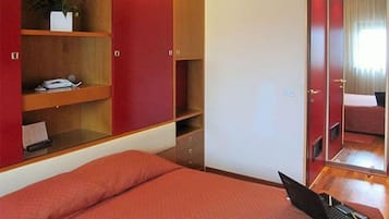 Quarto de hotel com uma cama, um laptop sobre a cama, uma parede vermelha, uma estante de madeira com uma planta e um espelho refletindo o quarto.