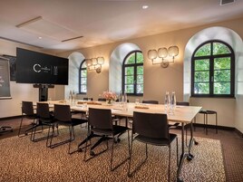 Sala de conferências com uma mesa comprida, cadeiras, tela de projetor e janelas em arco com luz natural.