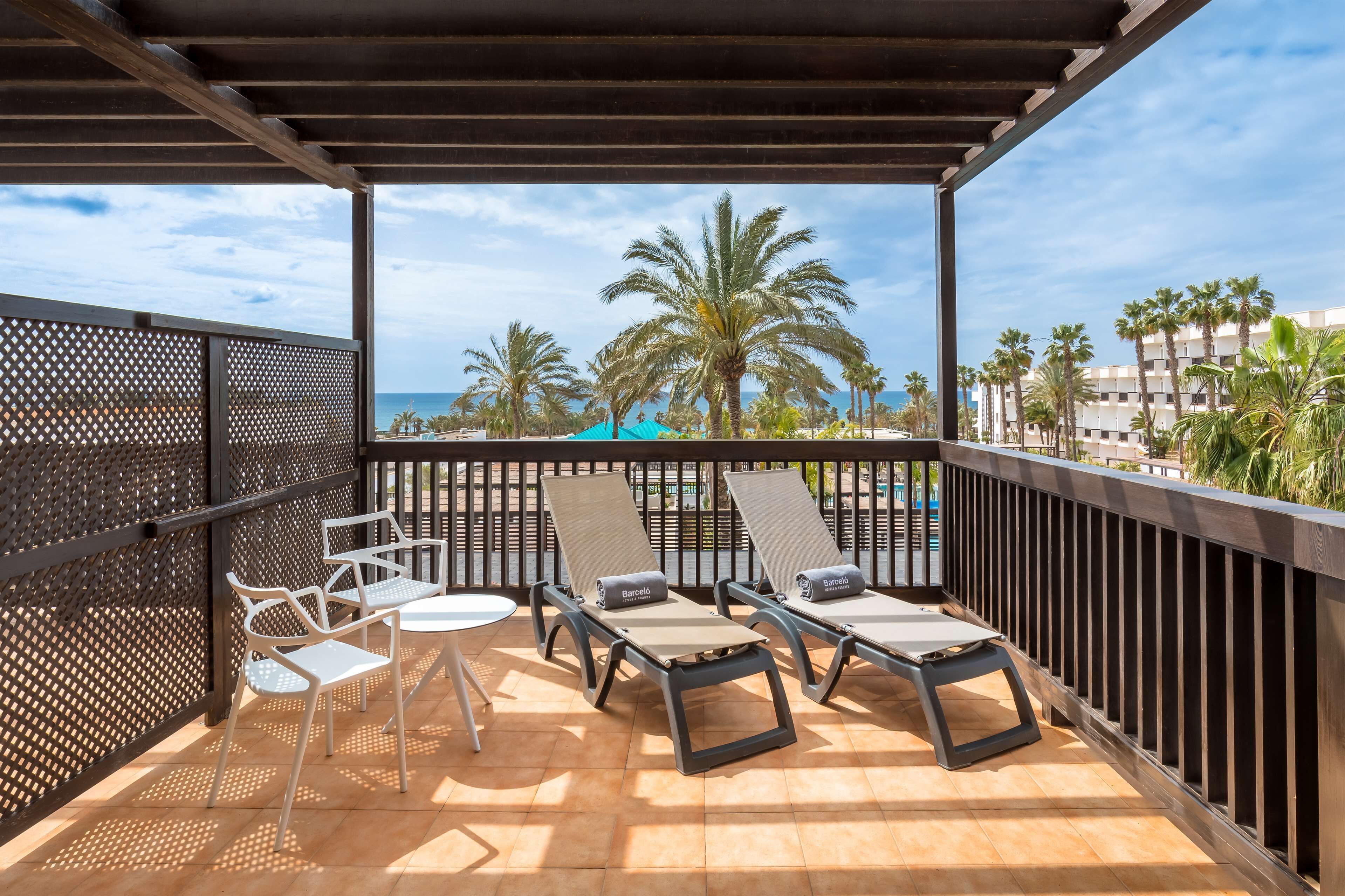 Área externa coberta com cadeiras de estar e uma mesa, com vista para uma piscina e palmeiras.