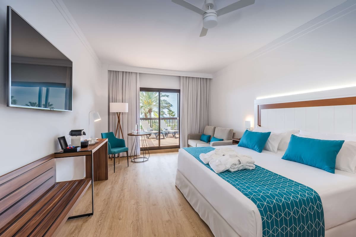 Quarto de hotel com cama, escrivaninha, cadeira, sofá, varanda e ventoinha no teto.