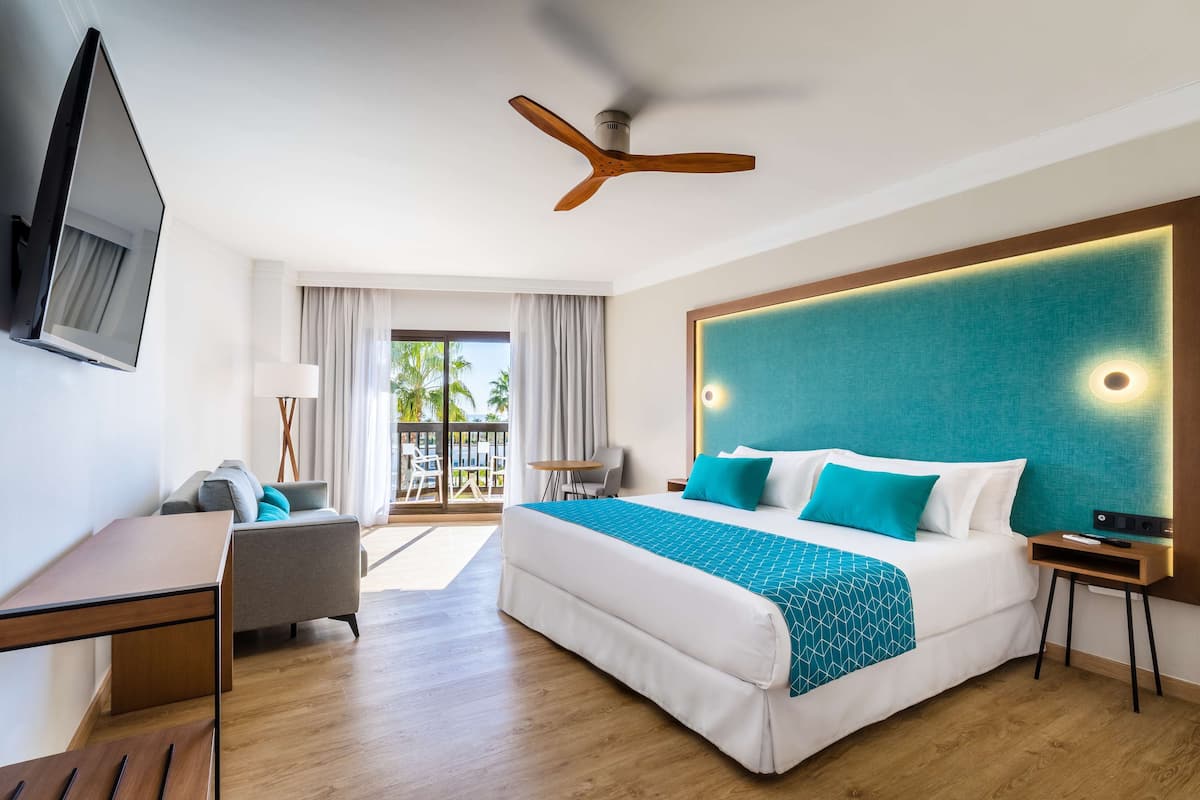 Quarto de hotel moderno com uma cama grande, uma escrivaninha, uma cadeira, uma TV, um ventilador de teto e uma varanda com vista para palmeiras.