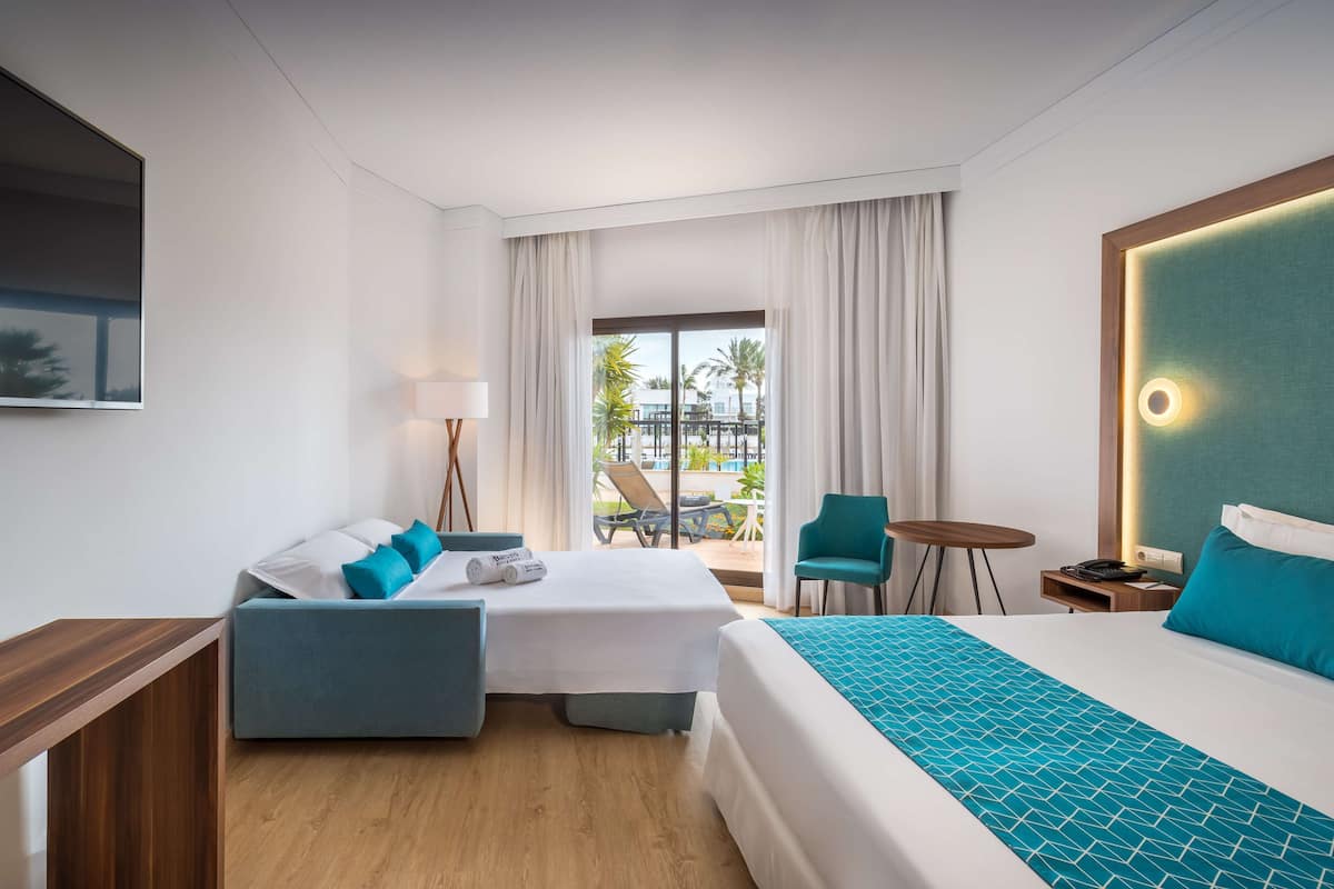 Quarto de hotel com uma cama grande, um sofá, uma escrivaninha e uma varanda com vista para palmeiras e uma piscina.