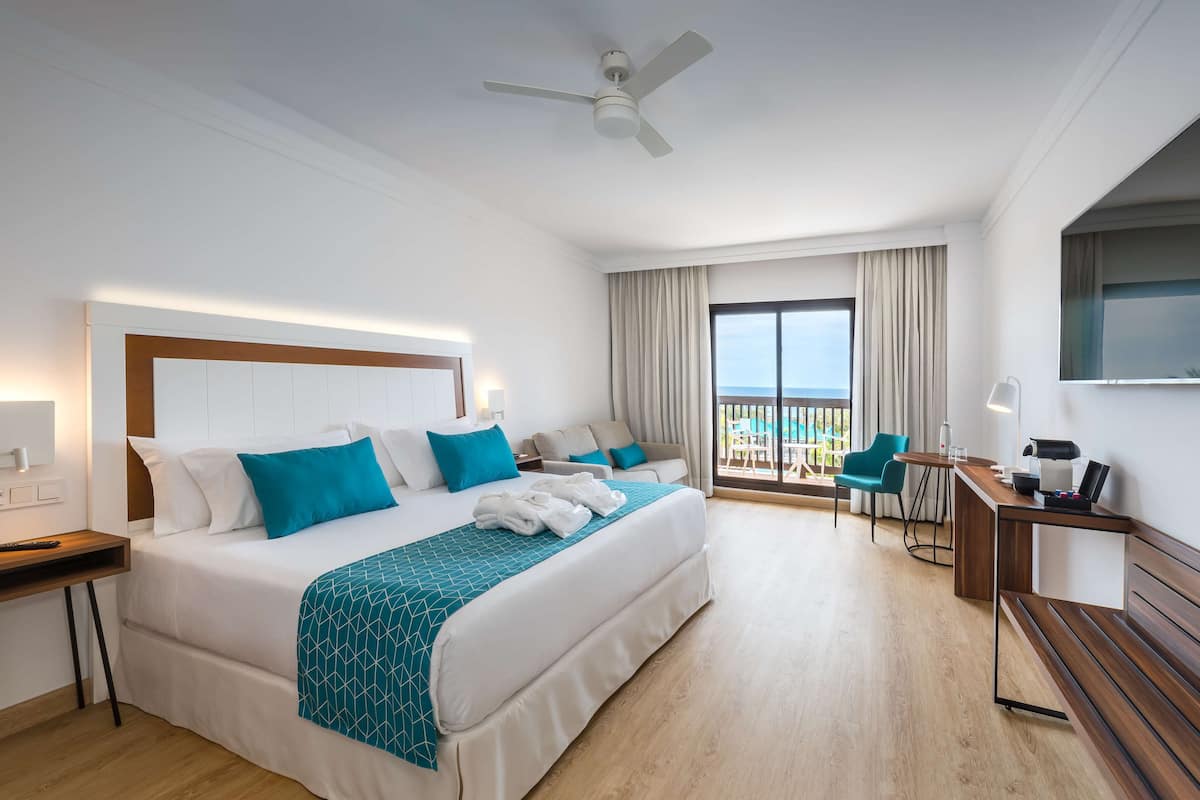 Quarto de hotel com uma cama grande, uma mesa com cadeira, varanda com vista e ventilador de teto.
