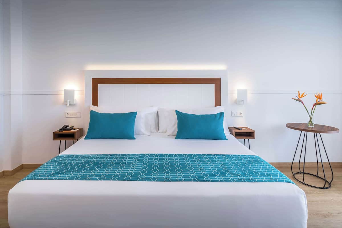 Quarto de hotel com uma cama grande, duas mesas de cabeceira e uma mesinha redonda com um vaso de flores.