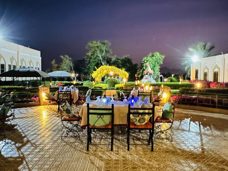 Uma área externa para refeições com mesas e cadeiras, uma fonte e um jardim bem iluminado à noite.