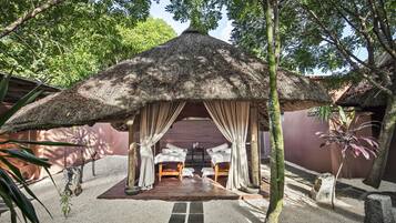 Um gazebo com telhado de palha, com duas cadeiras de madeira e um banco, rodeado por um caminho de cascalho e plantas tropicais.