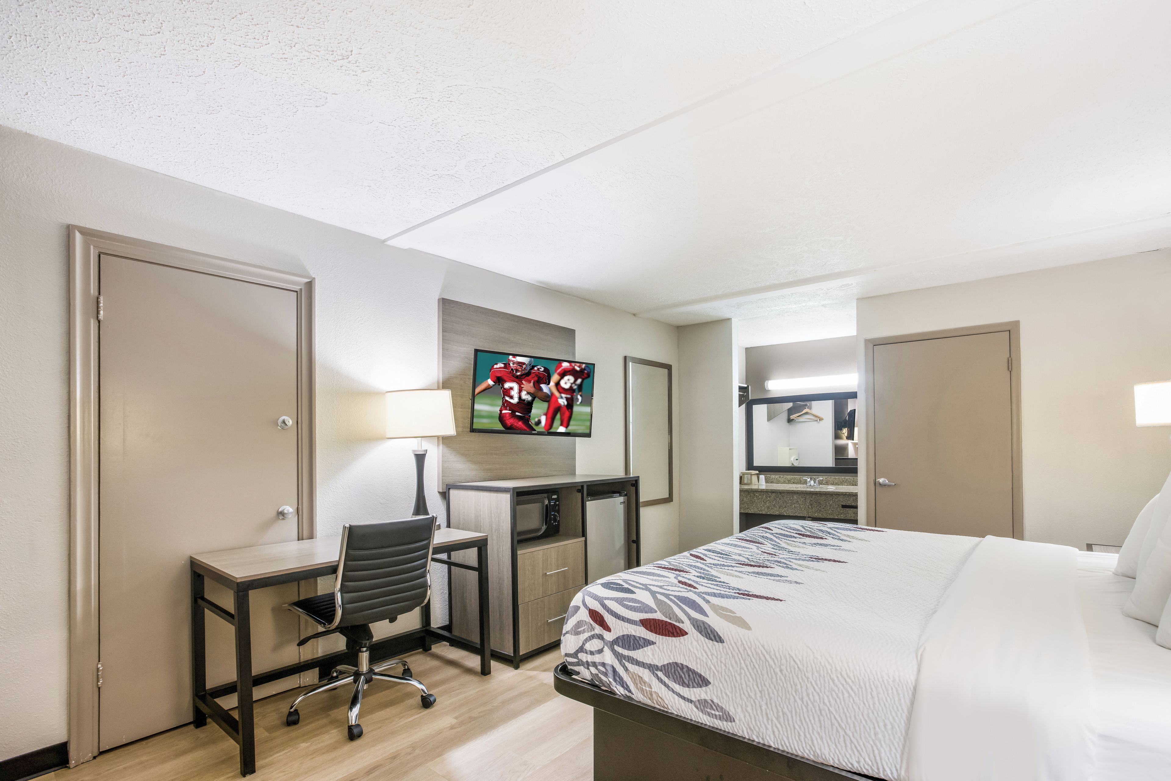 Quarto de hotel com cama, escrivaninha, cadeira, televisão e banheiro visível através de uma porta aberta.