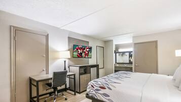 Quarto de hotel com cama, escrivaninha, cadeira, televisão e banheiro visível através de uma porta aberta.