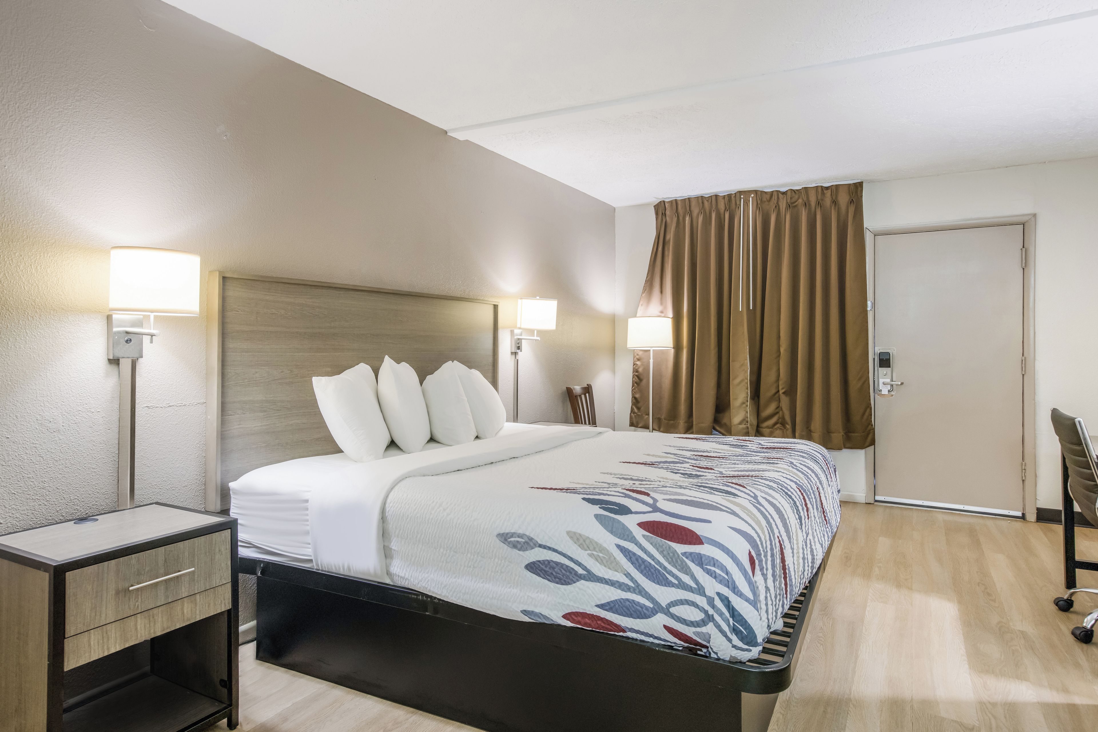 Quarto de hotel com cama, abajures, uma escrivaninha e uma porta.