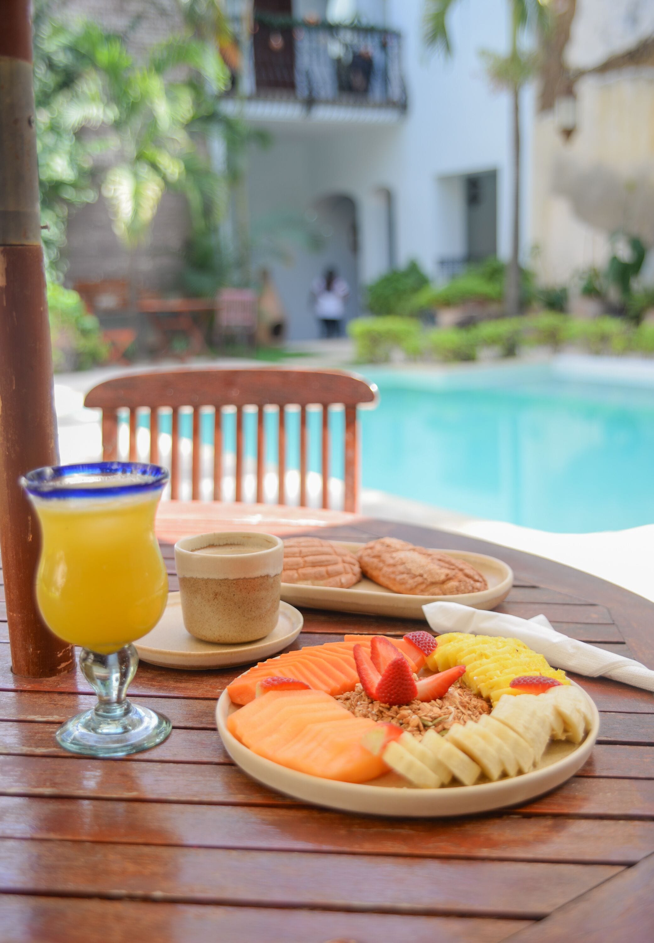 Uma mesa de madeira com uma travessa de frutas, um copo de suco de laranja e uma xícara de café, com vista para a piscina.