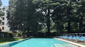 Piscina com água azul cristalina, cercada por árvores e uma fileira de espreguiçadeiras azuis e brancas.