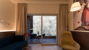 Uma sala de estar moderna com um sofá azul, uma poltrona amarela, uma mesa de centro de madeira e uma janela grande com vista para árvores.