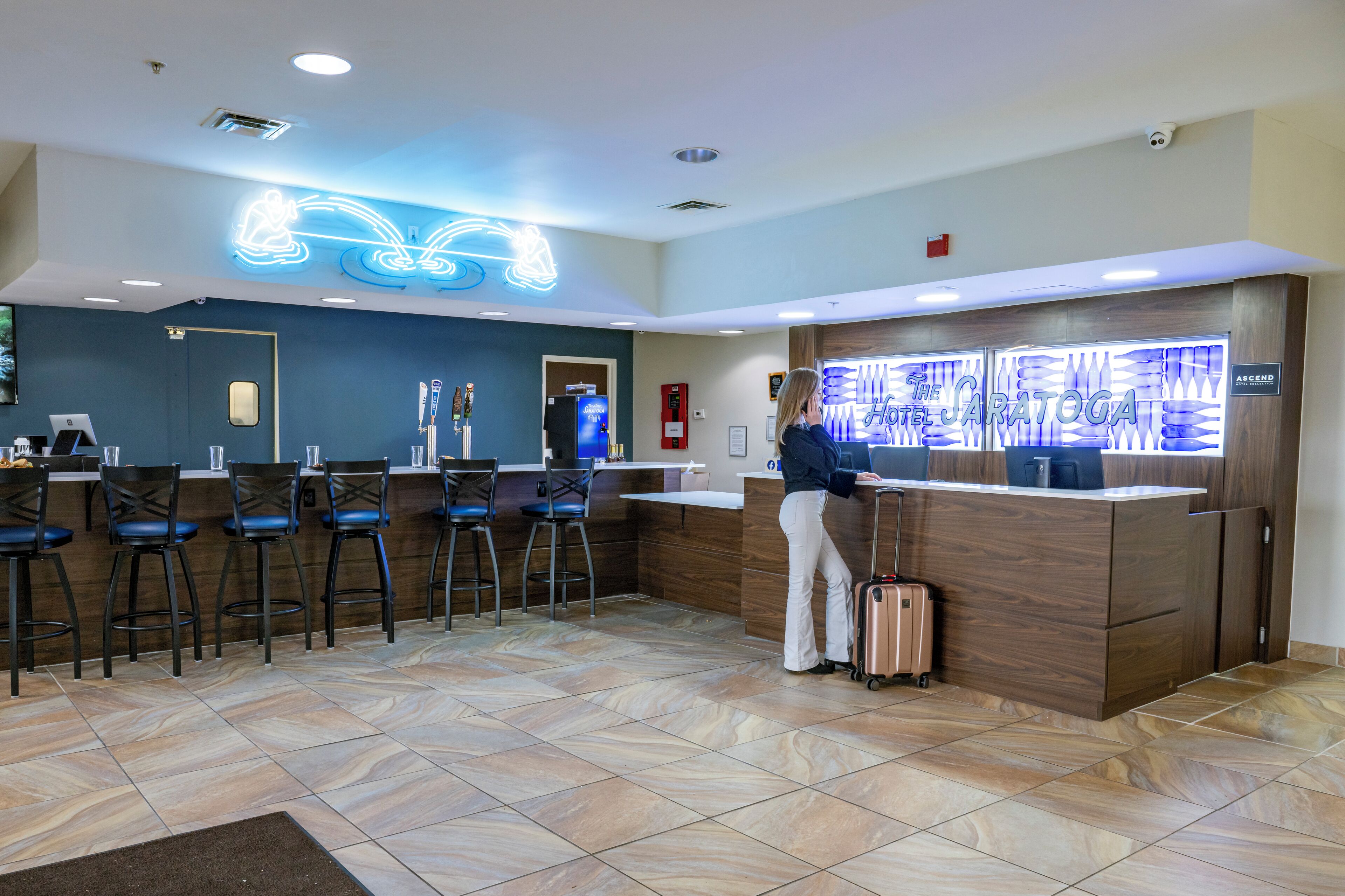 Uma área de recepção de hotel com um bar, banquetas, balcão de recepção e um letreiro de neon.