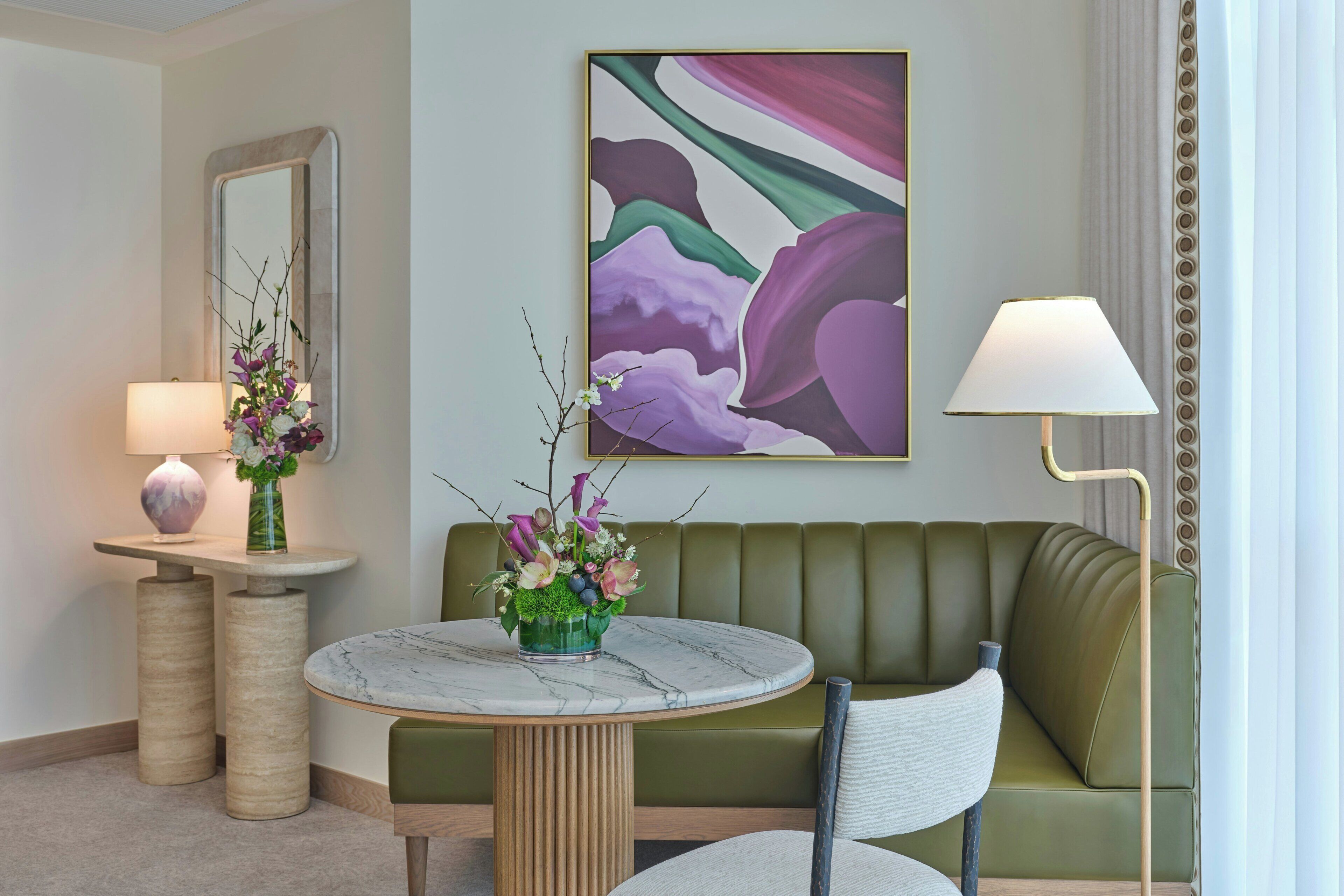 Um quarto com uma mesa redonda de mármore, um sofá, um abajur, um espelho e uma obra de arte floral na parede.
