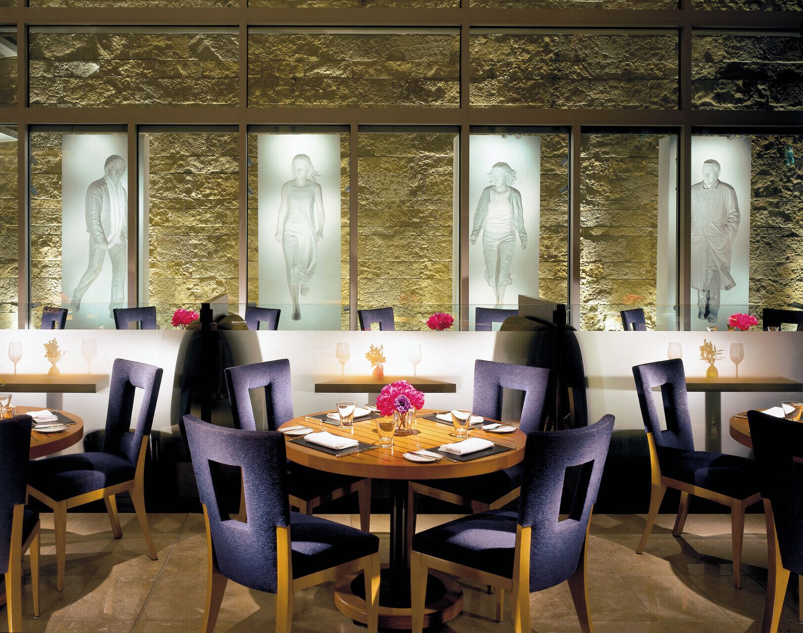 Restaurante com mesas redondas e cadeiras azuis, grandes janelas com painéis de vidro fosco e um fundo de parede de pedra.