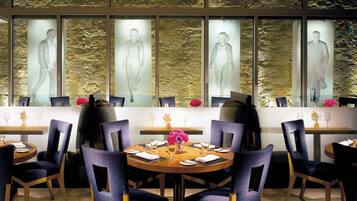 Restaurante com mesas redondas e cadeiras azuis, grandes janelas com painéis de vidro fosco e um fundo de parede de pedra.