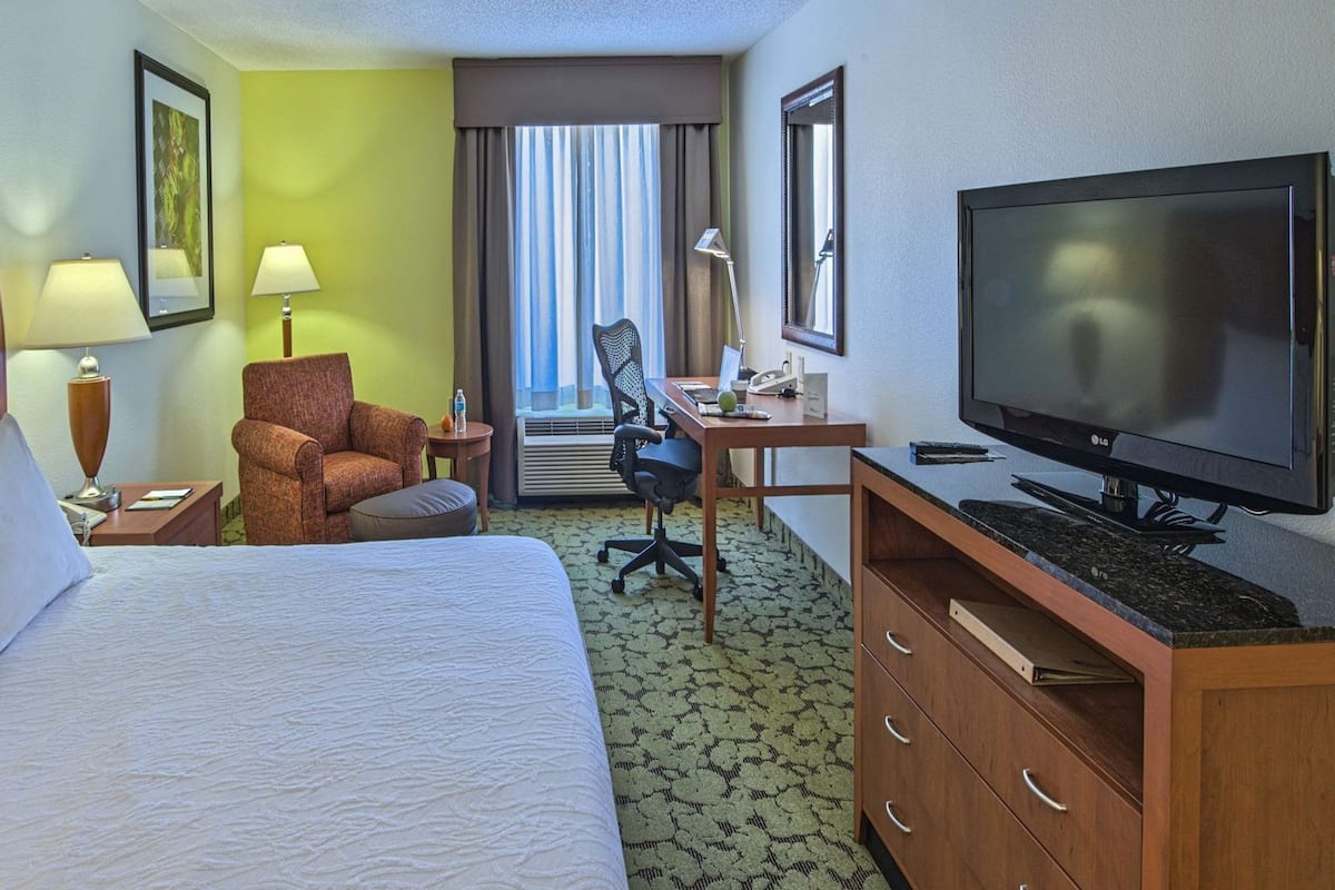 Quarto de hotel com cama, escrivaninha, cadeira, televisão e janela com cortinas.
