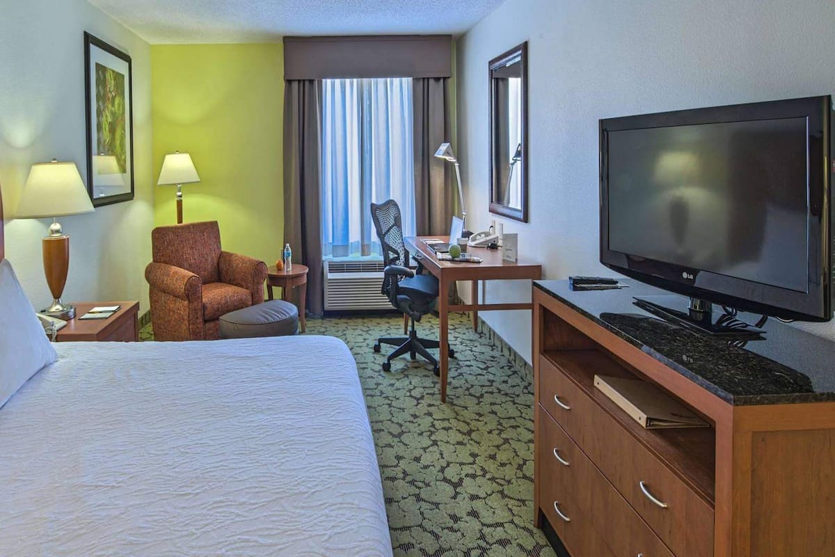 Quarto de hotel com cama, escrivaninha, cadeira, televisão e janela com cortinas.