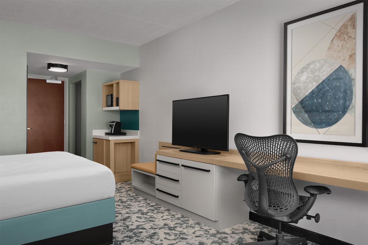 Quarto de hotel com cama, escrivaninha com computador, cadeira, máquina de café e uma obra de arte emoldurada na parede.