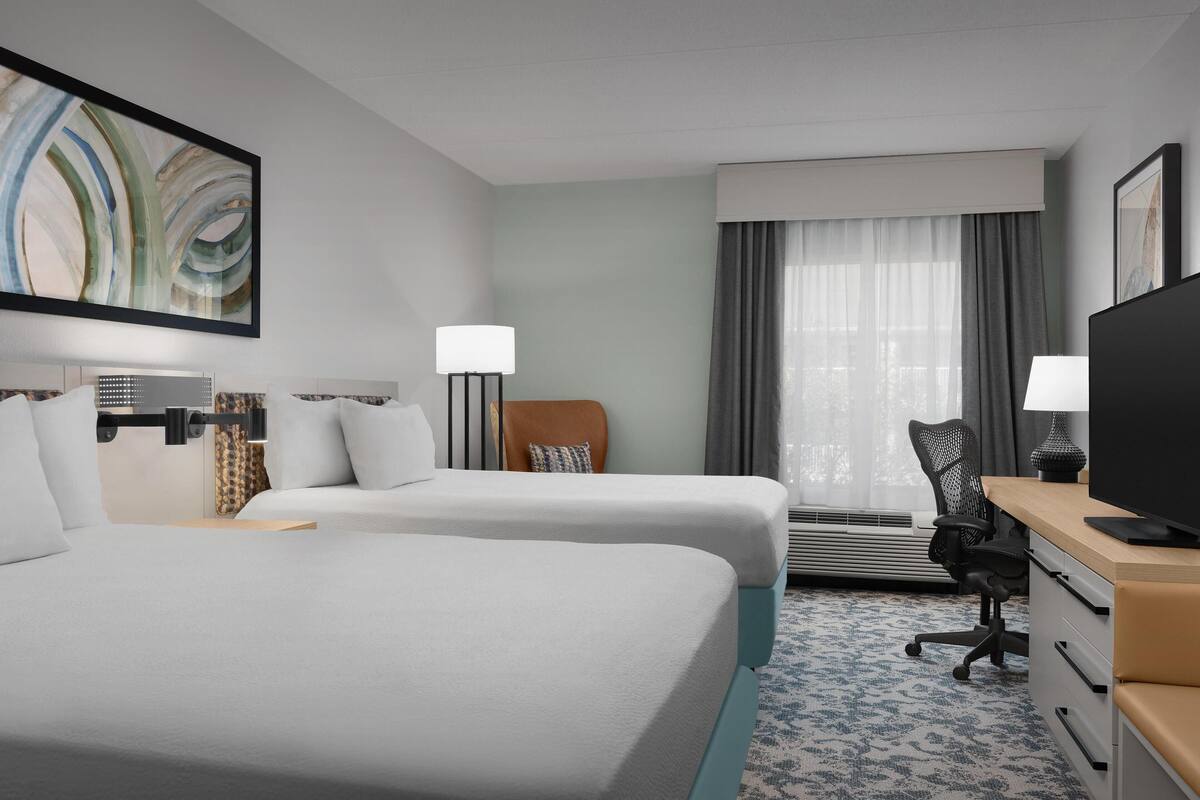 Quarto de hotel com duas camas, uma escrivaninha com computador, uma cadeira e uma janela grande com cortinas.