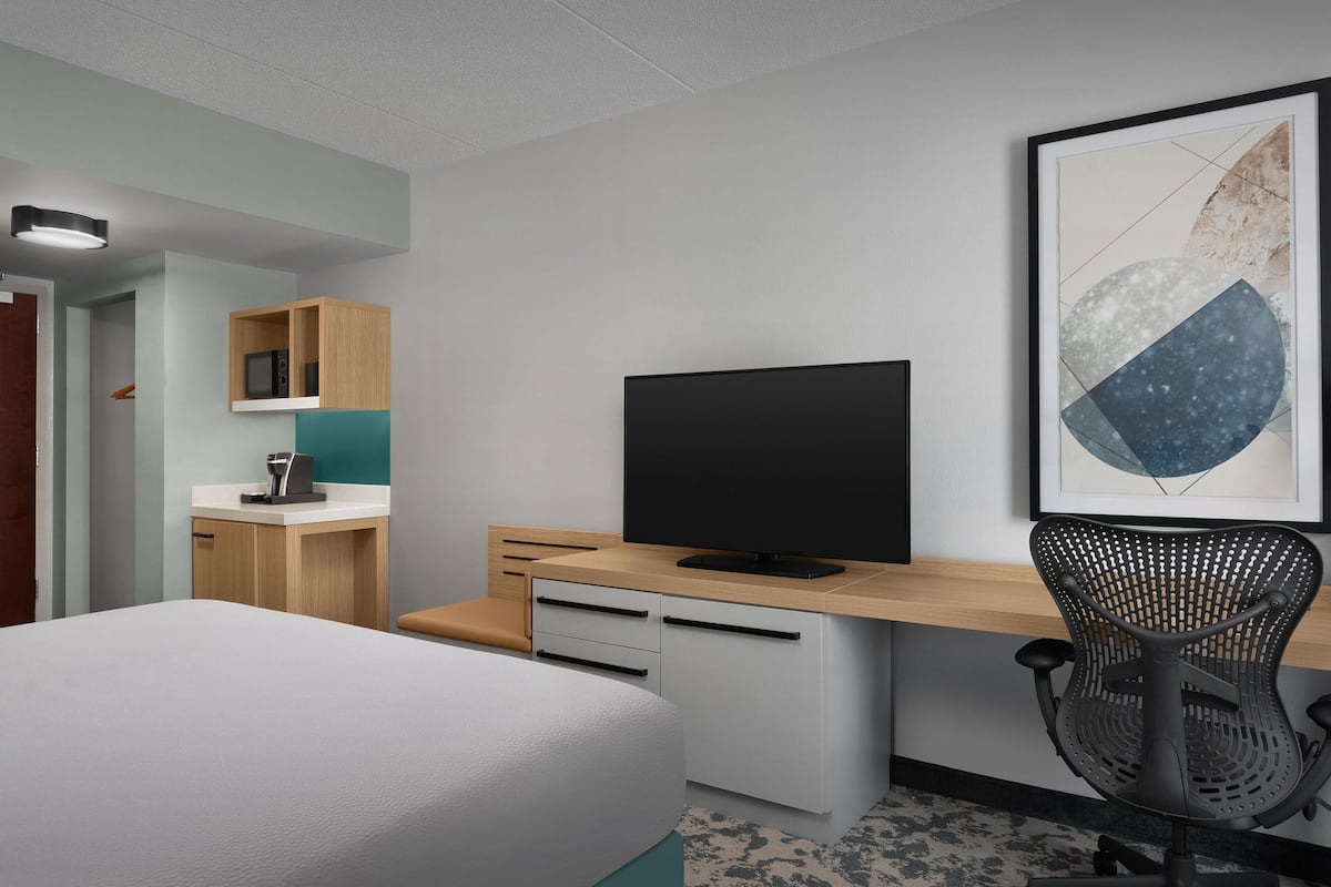Quarto de hotel moderno com cama, escrivaninha com computador e TV, uma cadeira e uma obra de arte abstrata emoldurada na parede.