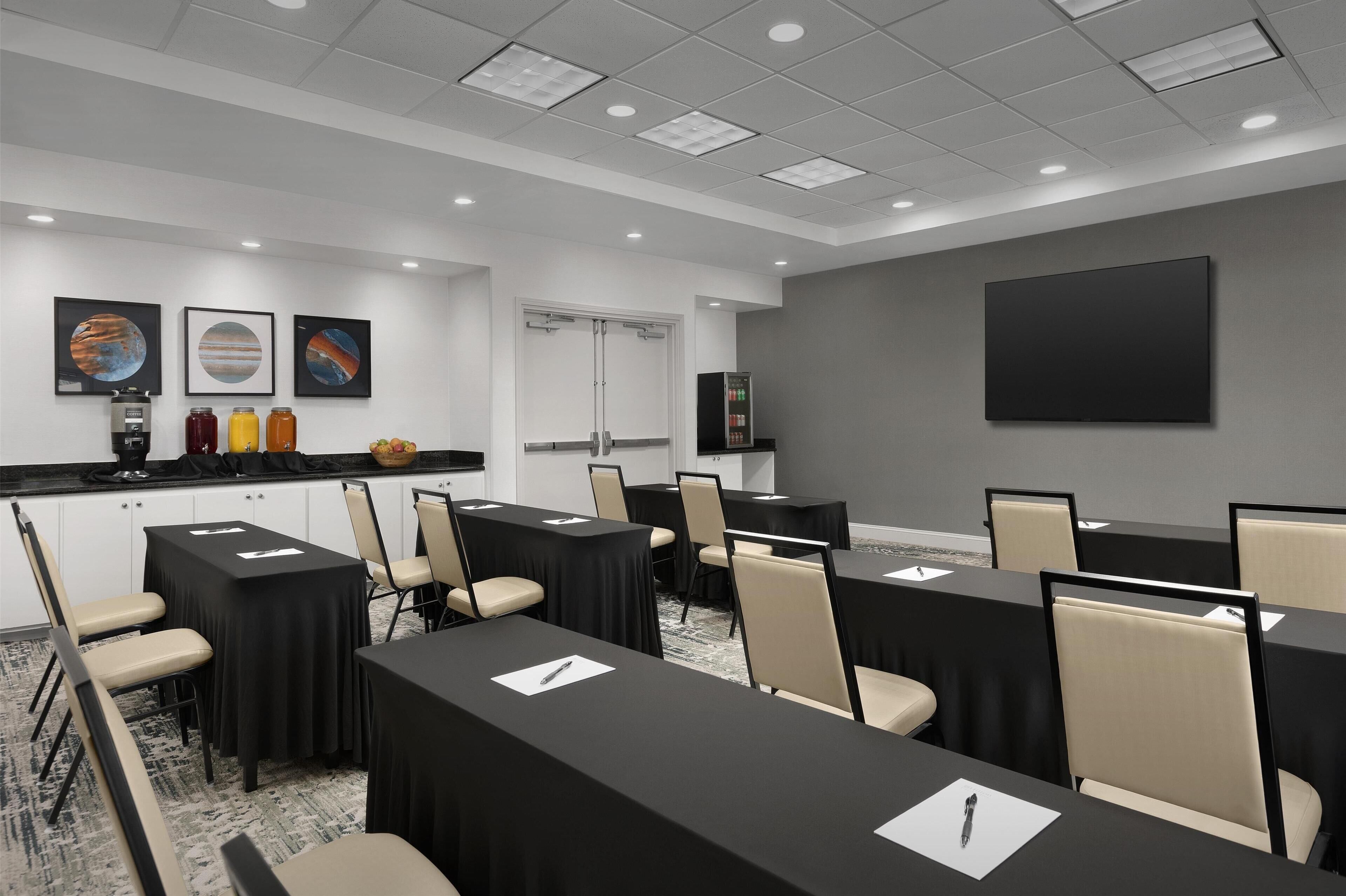 Uma sala de conferências com mesas, cadeiras, um espaço para café e uma tela grande na parede.