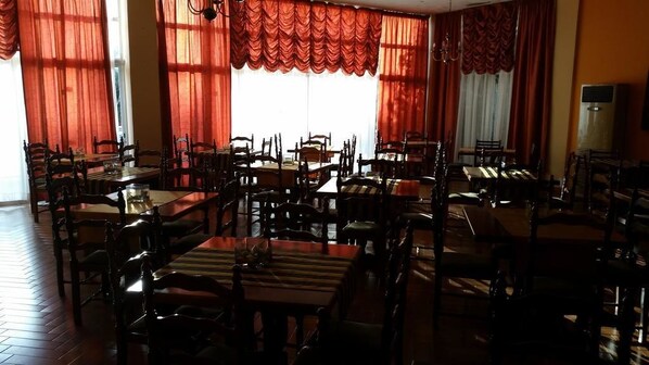 Uma sala de jantar com várias mesas e cadeiras, janelas amplas com cortinas vermelhas e um lustre.