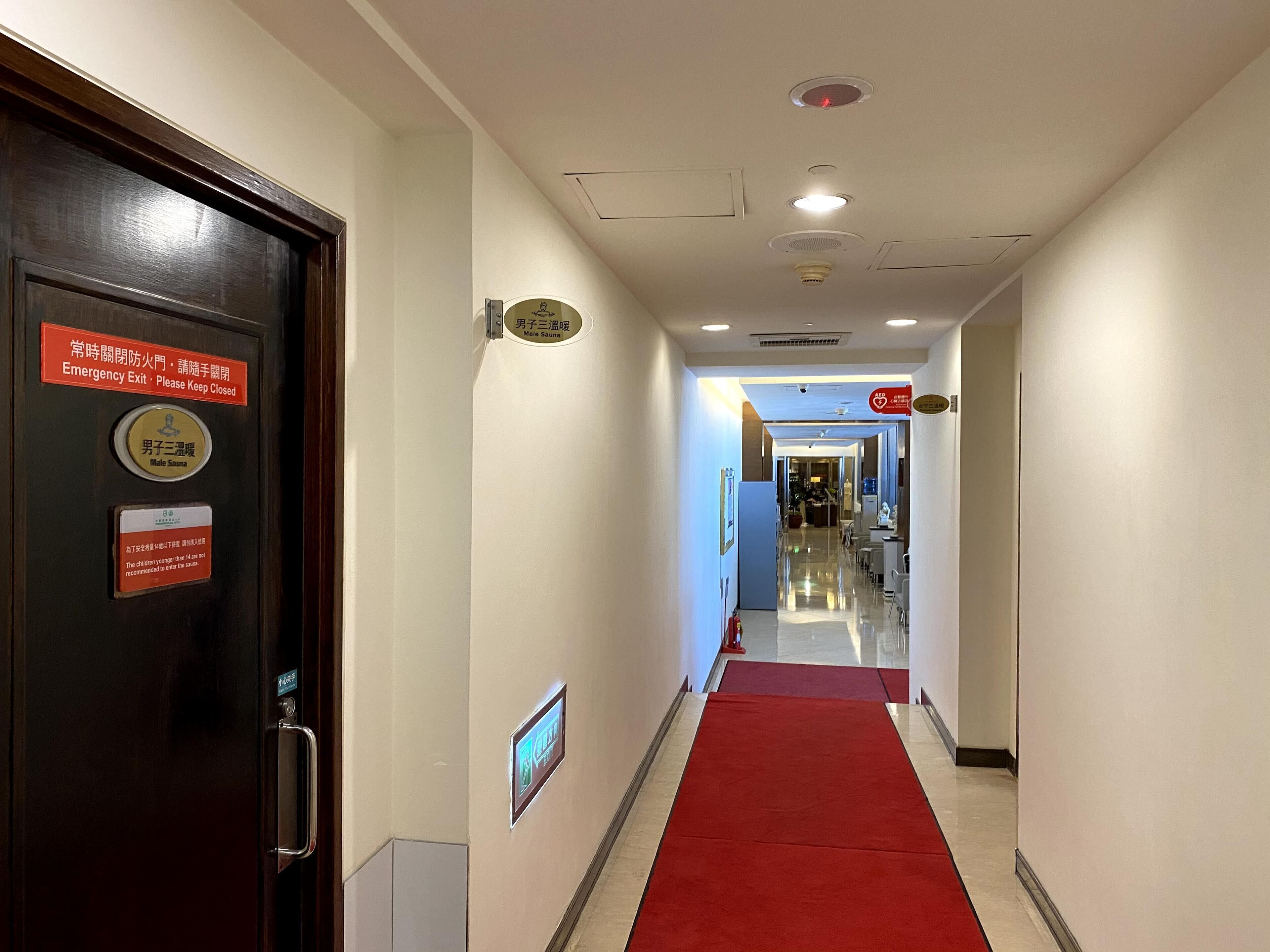 Corredor de hotel com tapete vermelho, placas de saída de emergência e sinalização para sauna masculina.