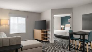Quarto de hotel com sofá, televisão sobre suporte de madeira, mesa de jantar com cadeiras, cama e tapete estampado.