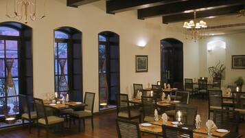 Restaurante com várias mesas de jantar dispostas com pratos, copos e velas. Janelas amplas com arcos, piso de madeira e tetos altos com vigas aparentes.